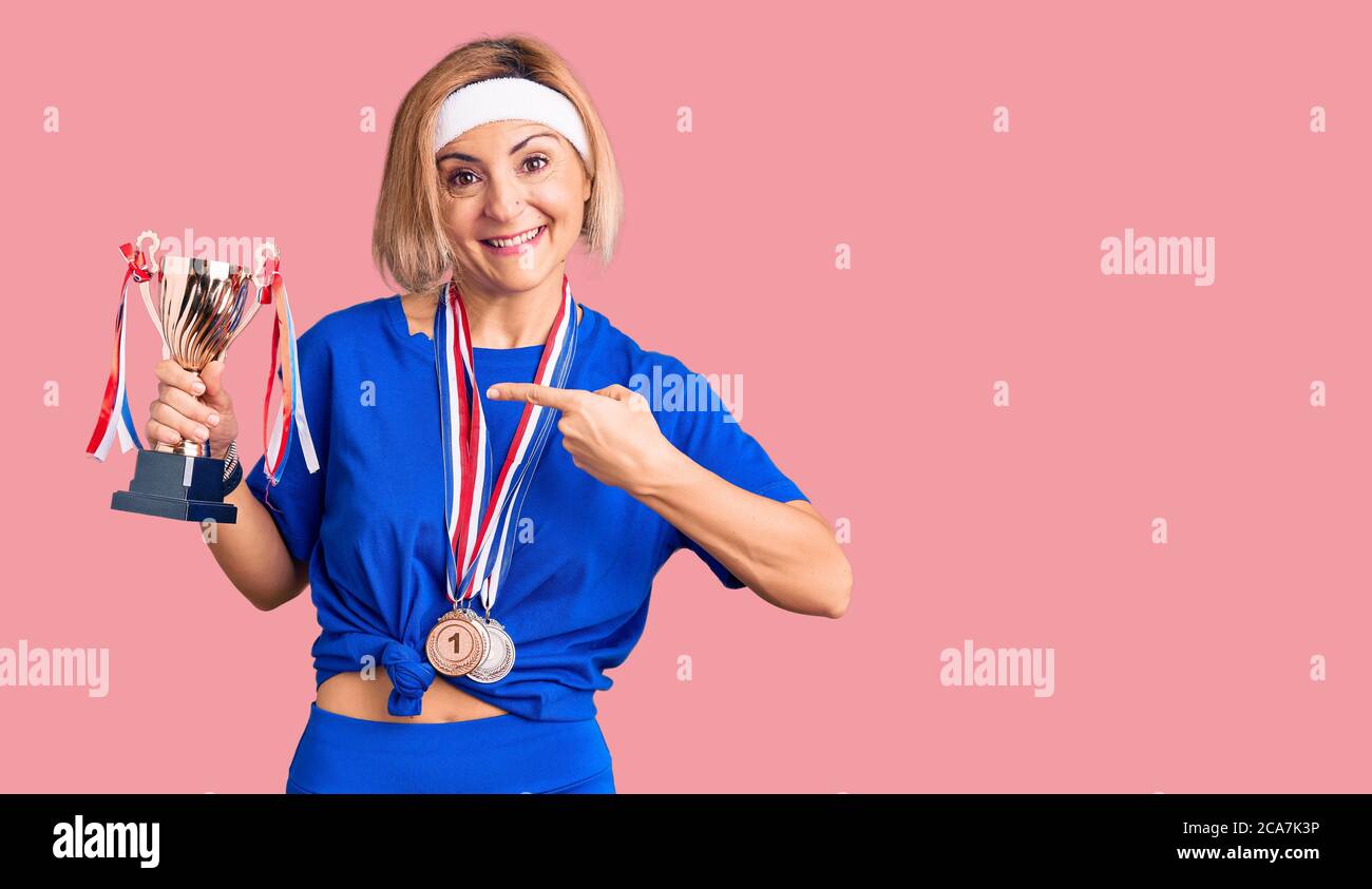 Young blonde woman holding champion trophy wearing medals smiling happy ...