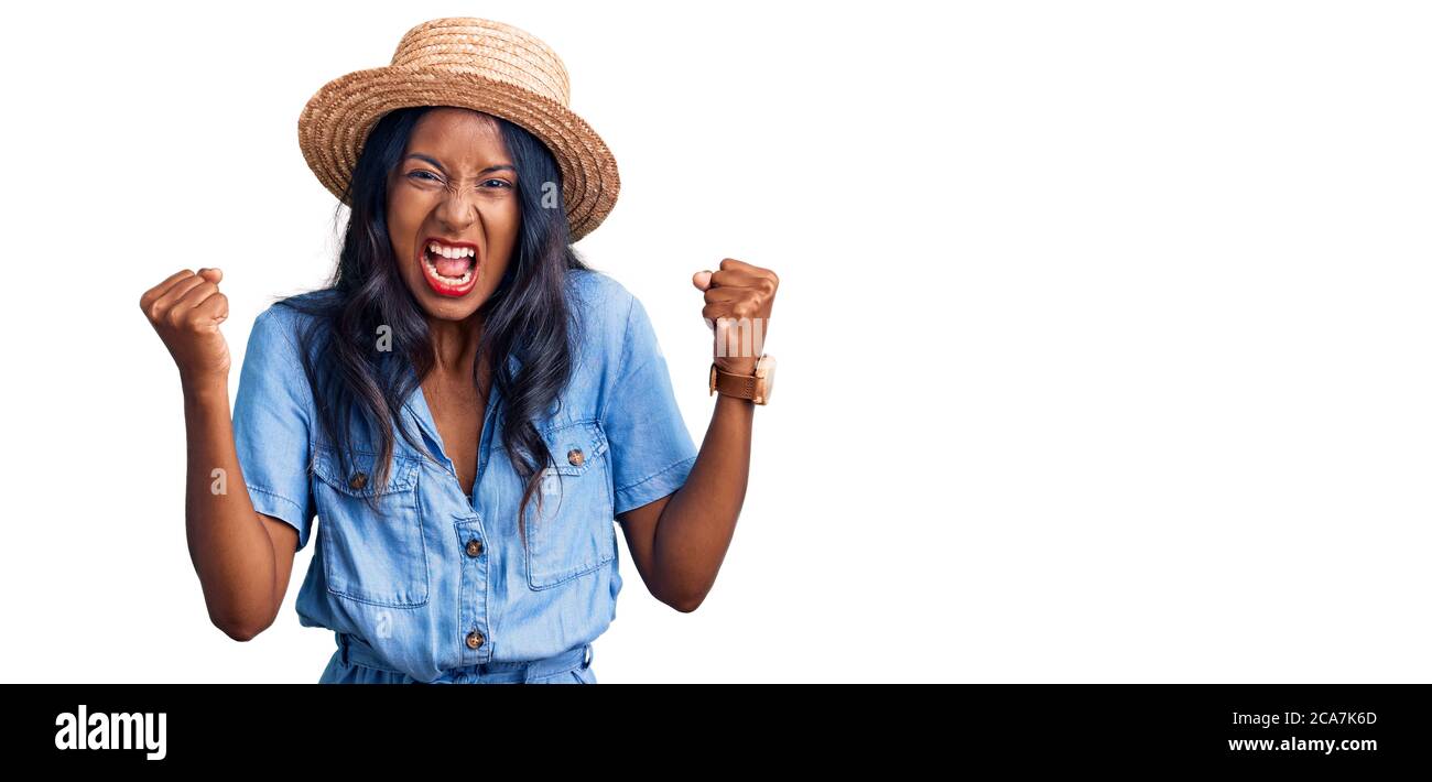 Young indian girl wearing summer hat angry and mad raising fists ...