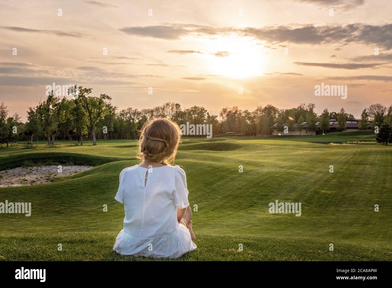 A lonely girl in a white dress sitting on golf course looking at sunset ...
