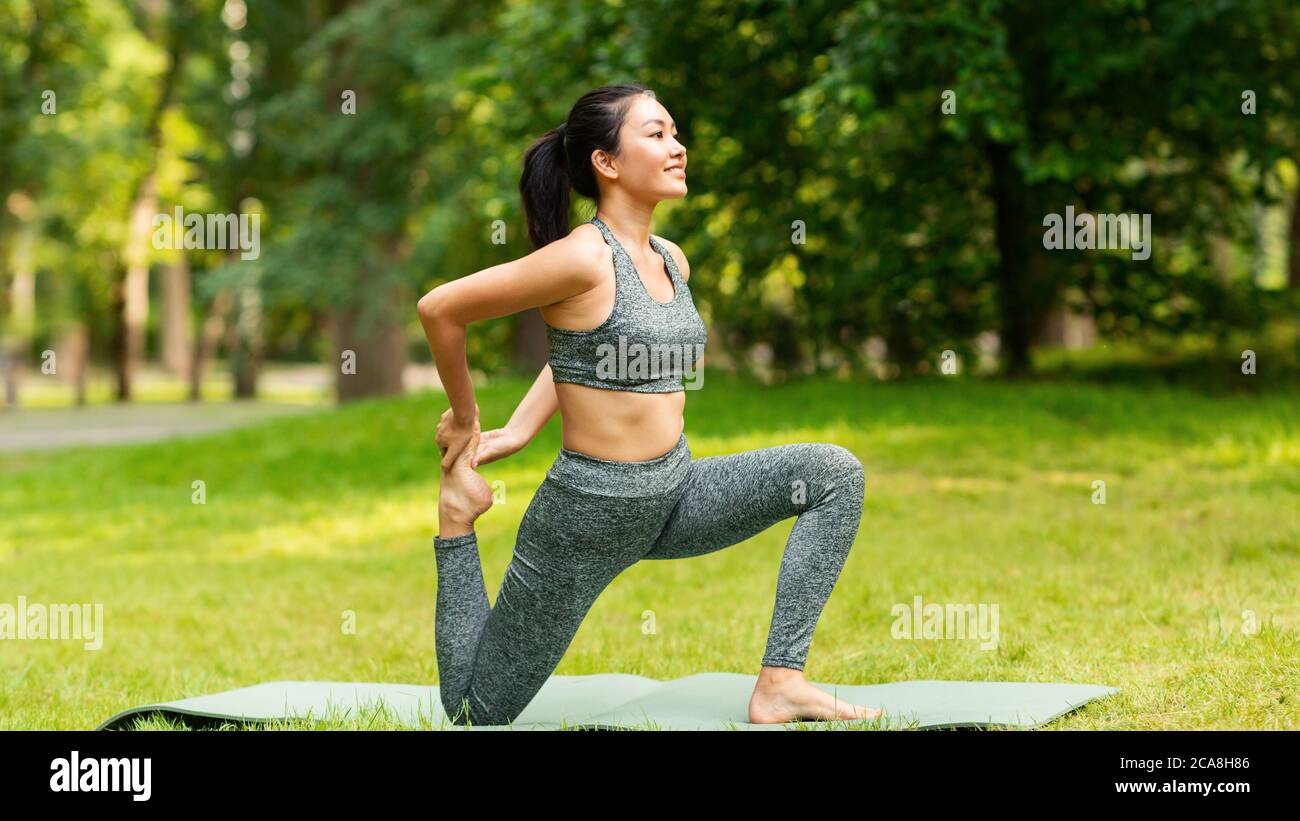 Sporty Asian girl doing yoga stretching exercises on mat at summer park ...