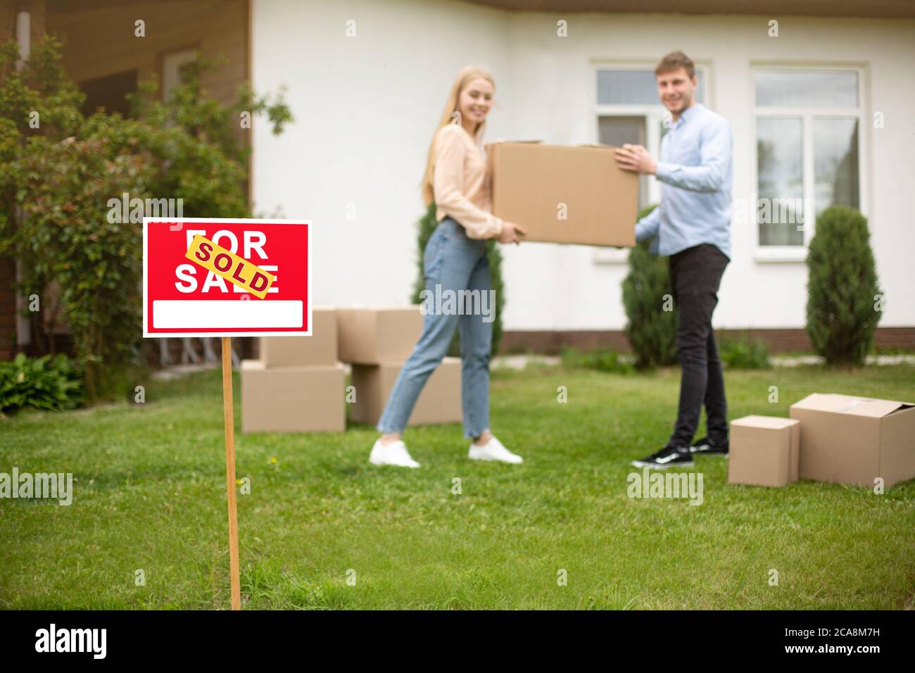 SOLD sign and young couple carrying cardboard box at house yard ...
