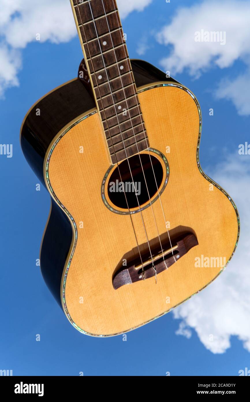 Acoustic baritone ukulele guitar held up to bright blue sky with fluffy ...