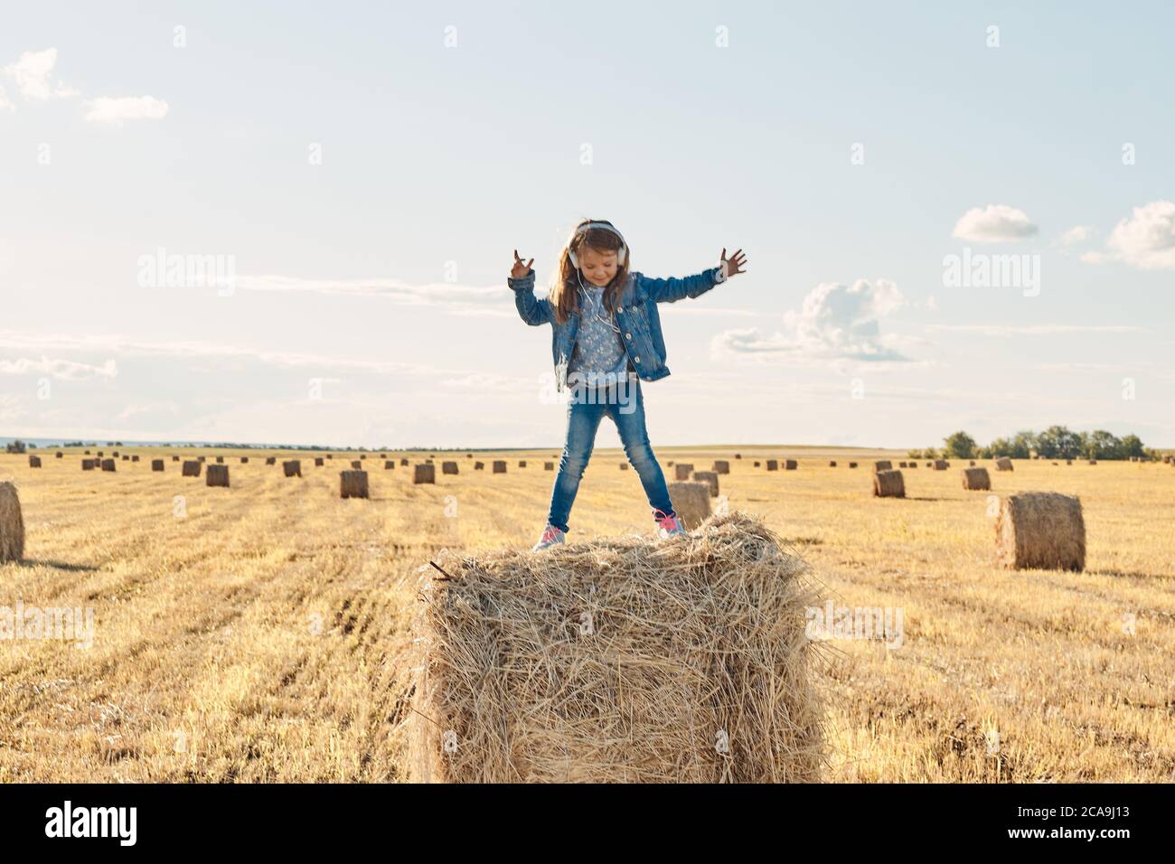 A cute little girl listens to music and dances on a haystack. High ...