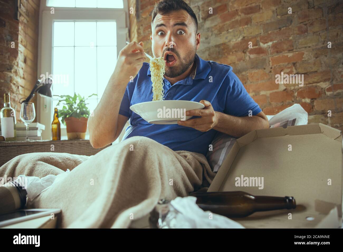 Shocked eating instant noodle. Lazy man living in his bed surrounded ...
