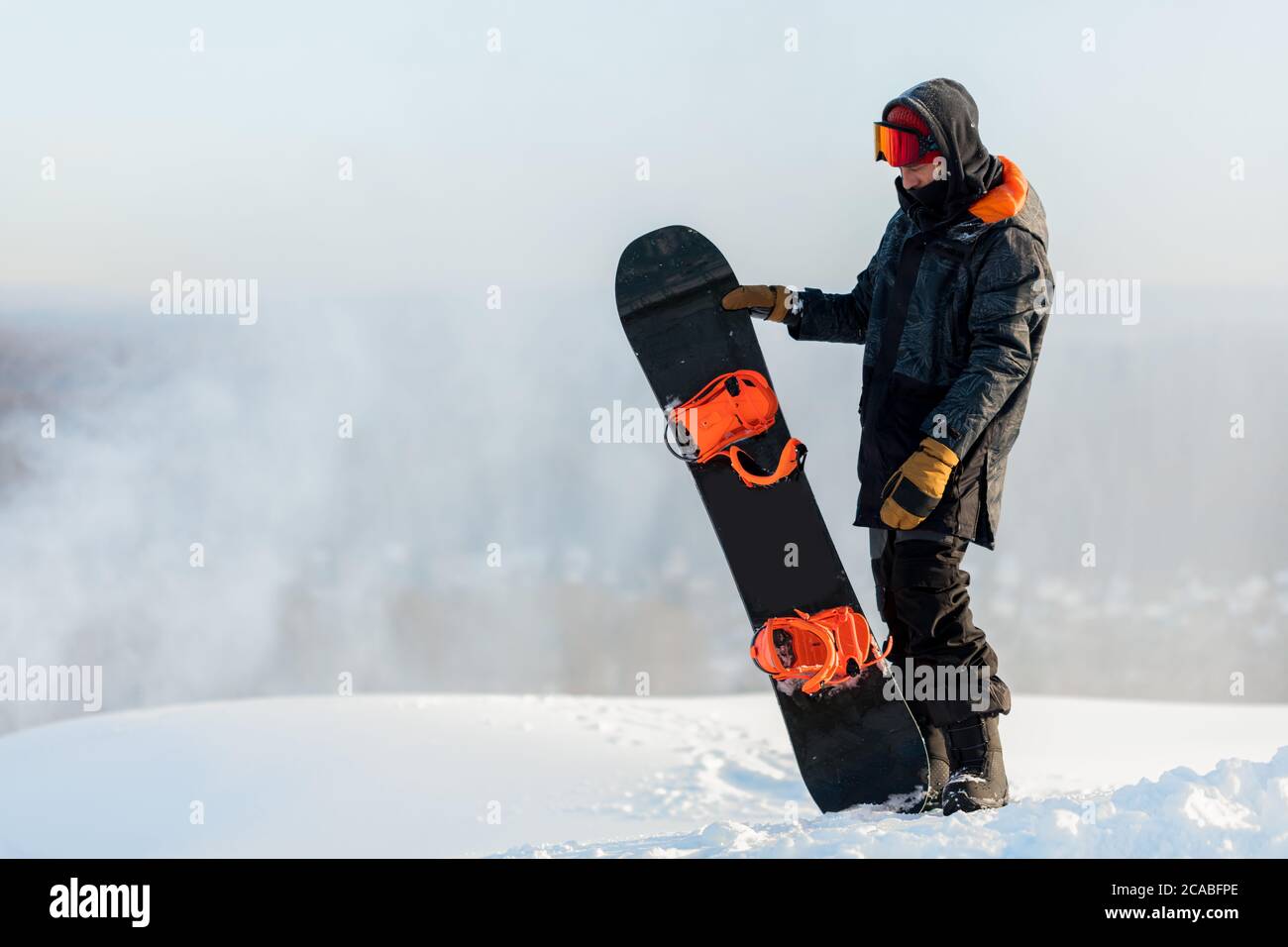 sporty man looking at snowboard while standing on the hill. side view ...