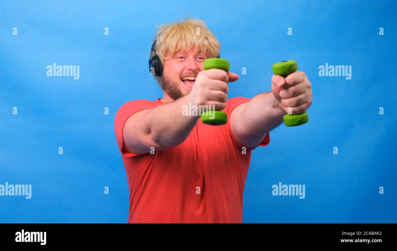 Funny fat man freaky in a wig with dumbbells doing exercises on a blue ...