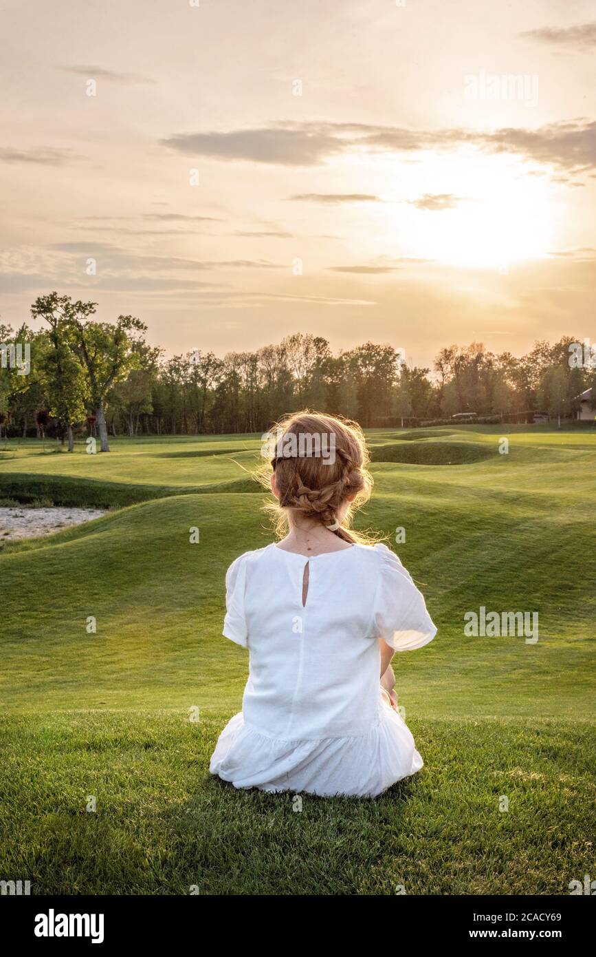 A lonely girl in a white dress sitting on golf course looking at sunset ...