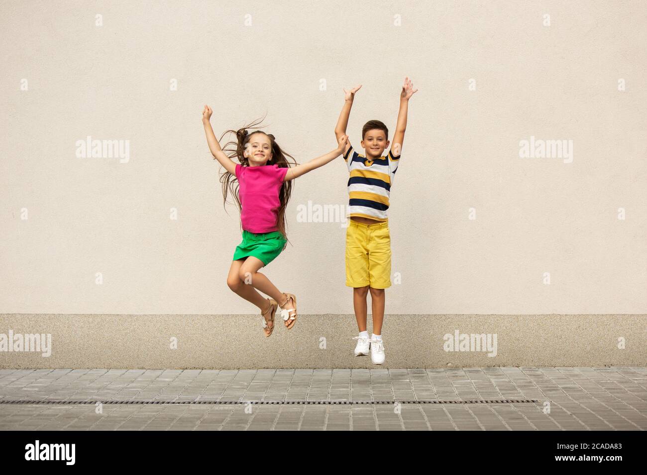 Two smiling kids, boy and girl jumping together in town, city in summer ...
