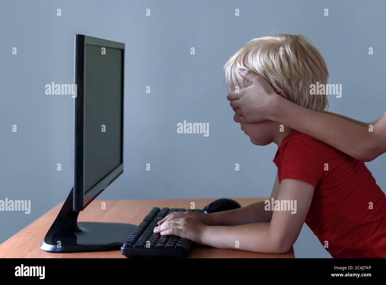 Children hands puts over eyes to a little boy who uses a computer ...