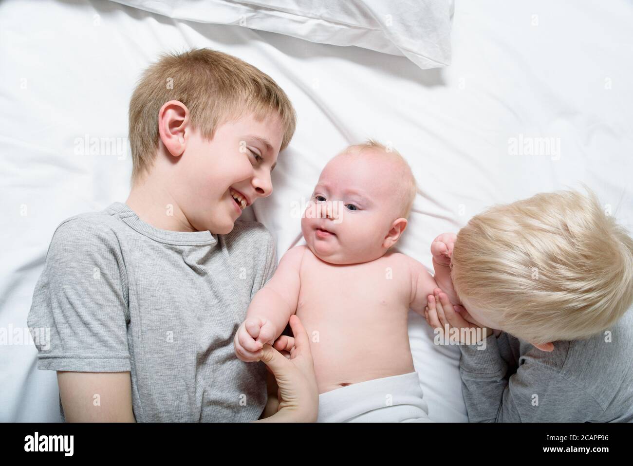 Two older brothers play and laugh with the youngest baby in a white bed ...