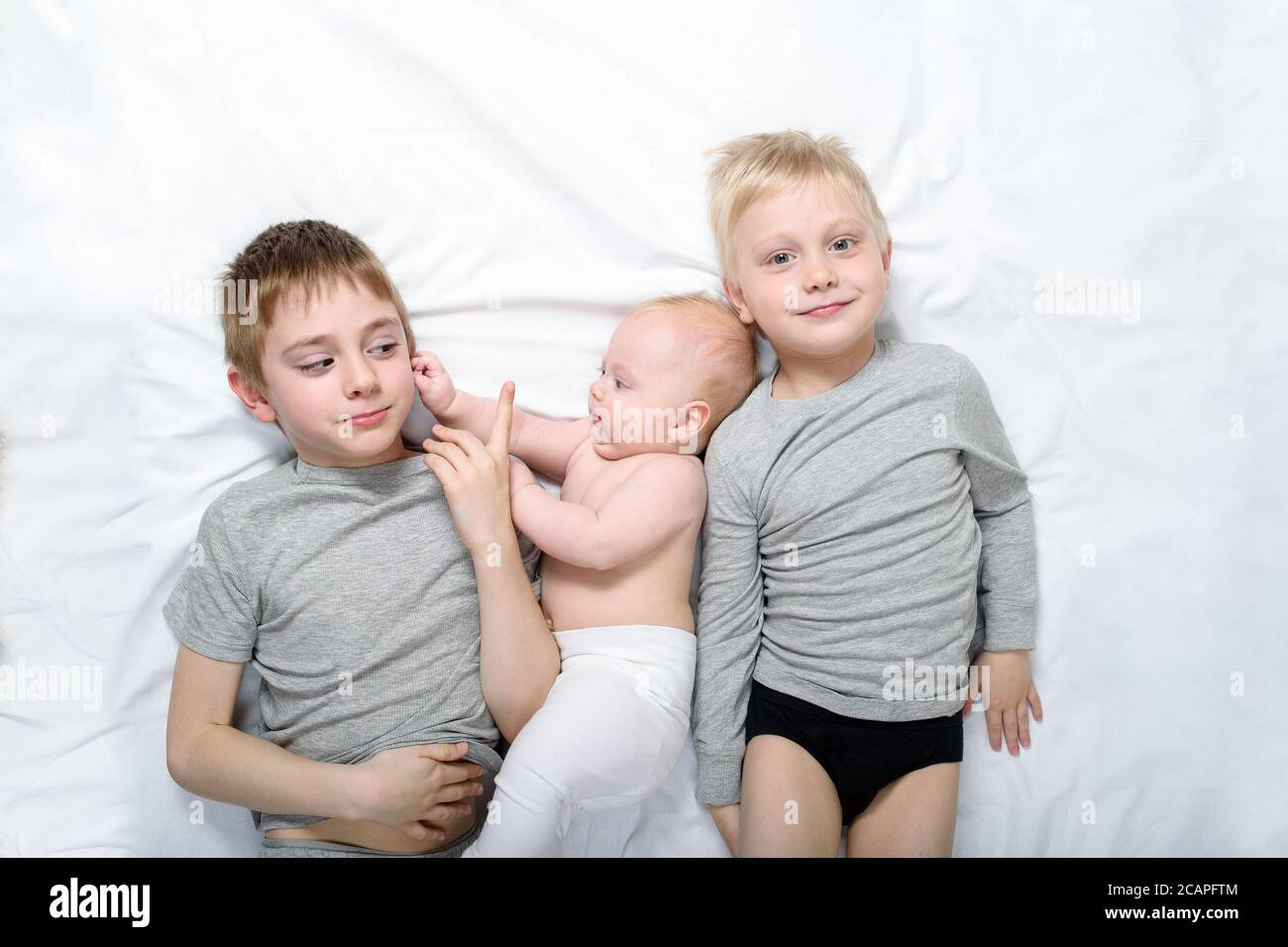 Two older brothers play and laugh with the youngest baby in a white bed ...