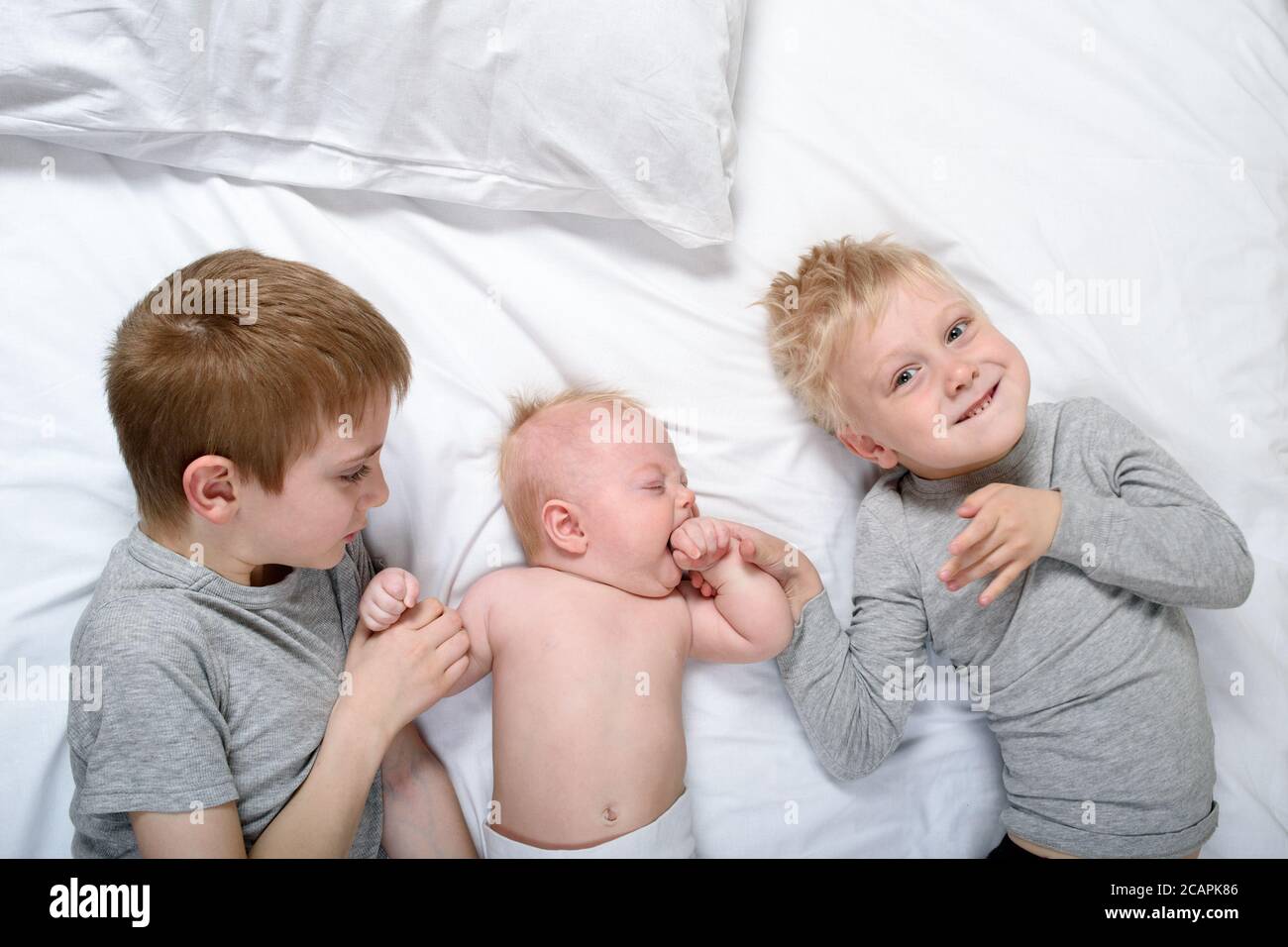Two older brothers play and laugh with the youngest baby in a white bed ...