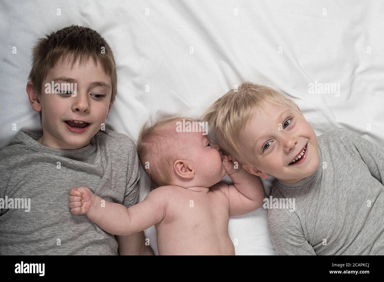 Two older brothers play and laugh with the youngest baby in a white bed ...