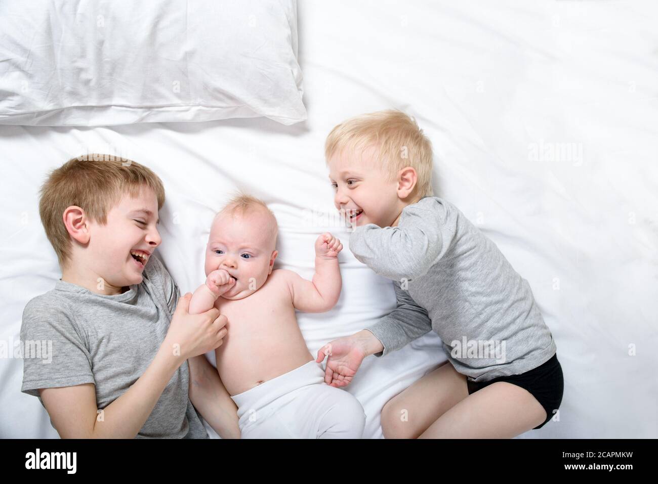 Two older brothers play and laugh with the youngest baby in a white bed ...