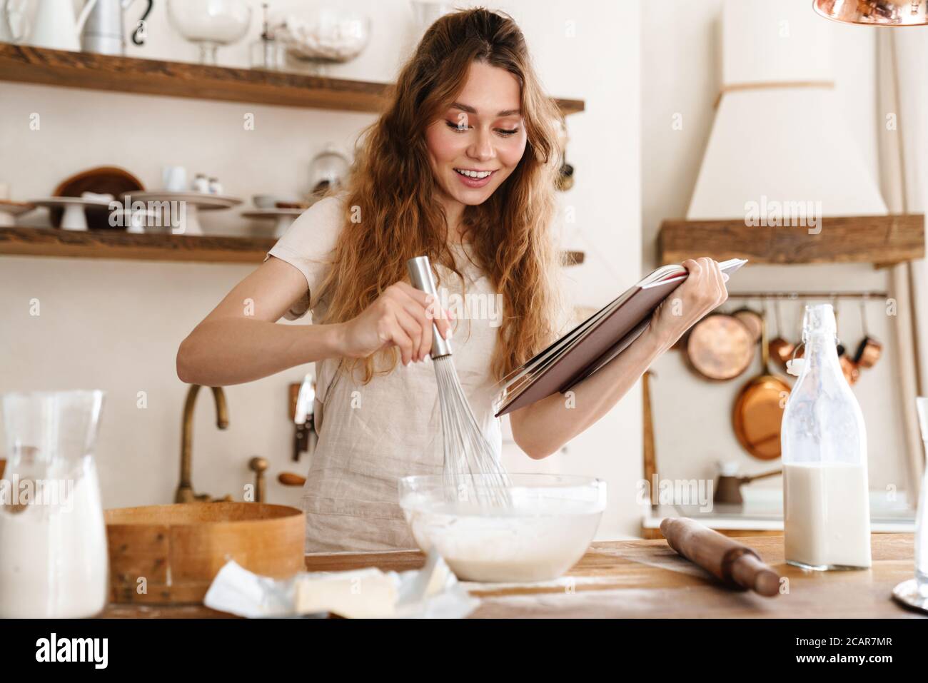 Attractive young cheerful girl baking at the kitchen, making dough ...