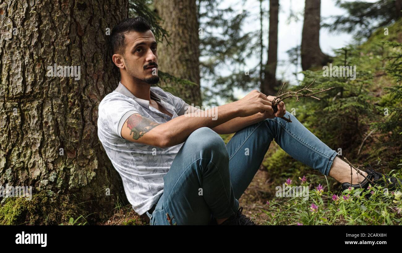 Handsome young man leaning on tree is dealing with wood stick in his ...