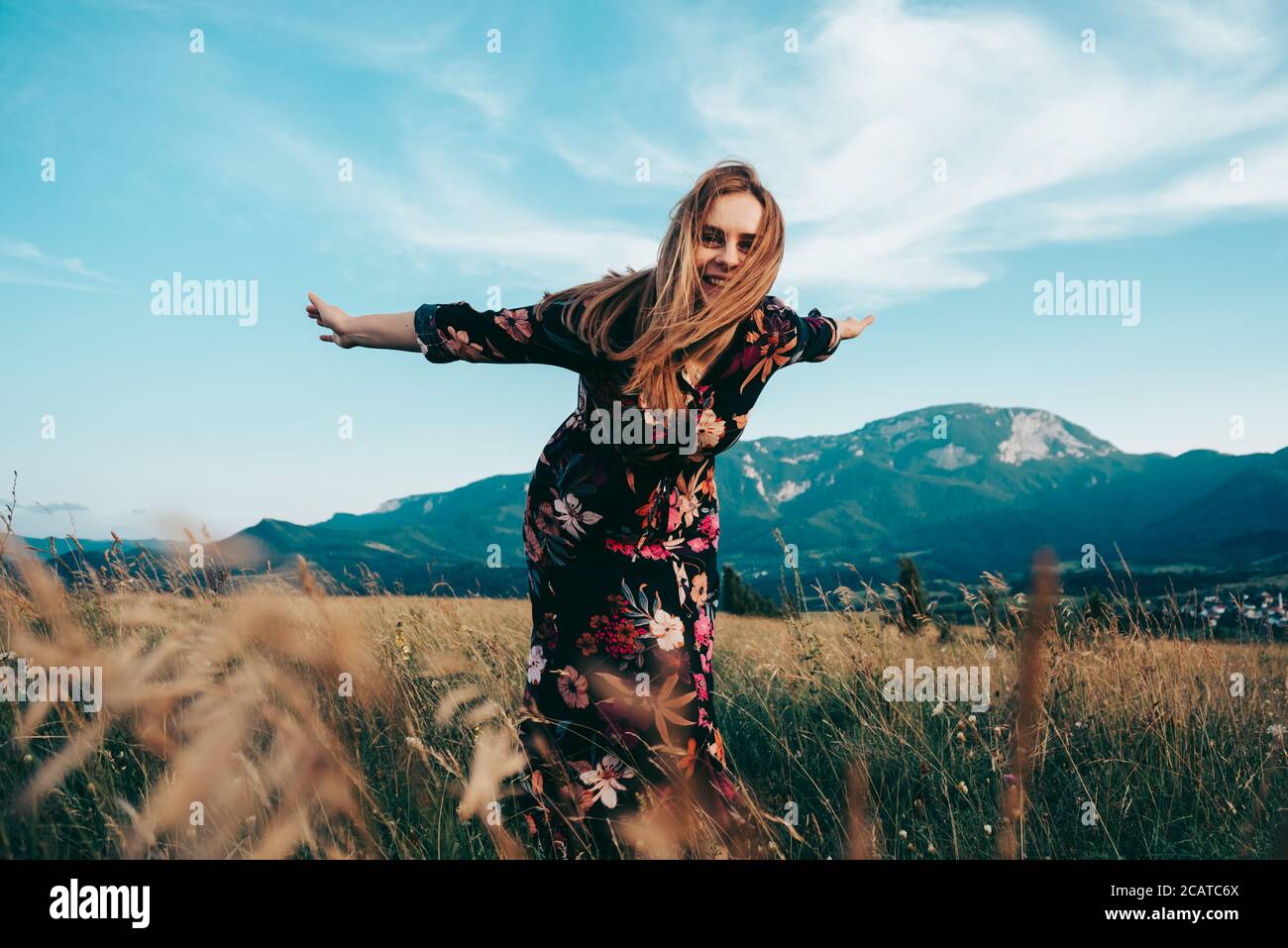 Happy woman sitting in grass field with mountains in background in ...