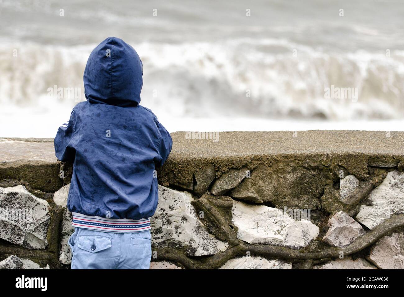 Boy in blue jacket with hood stands with his back against the pier ...