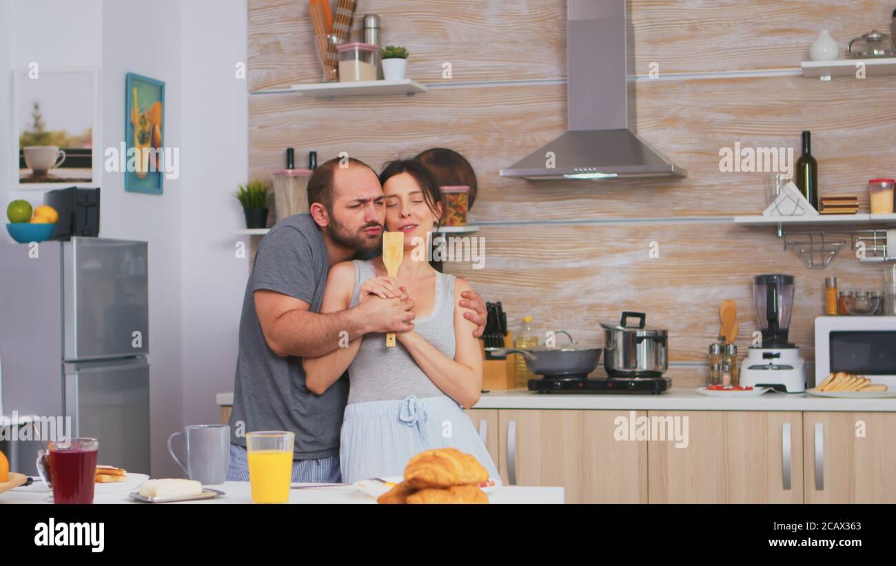 Wife singing on wooden spoon while dancing with husband in kitchen ...