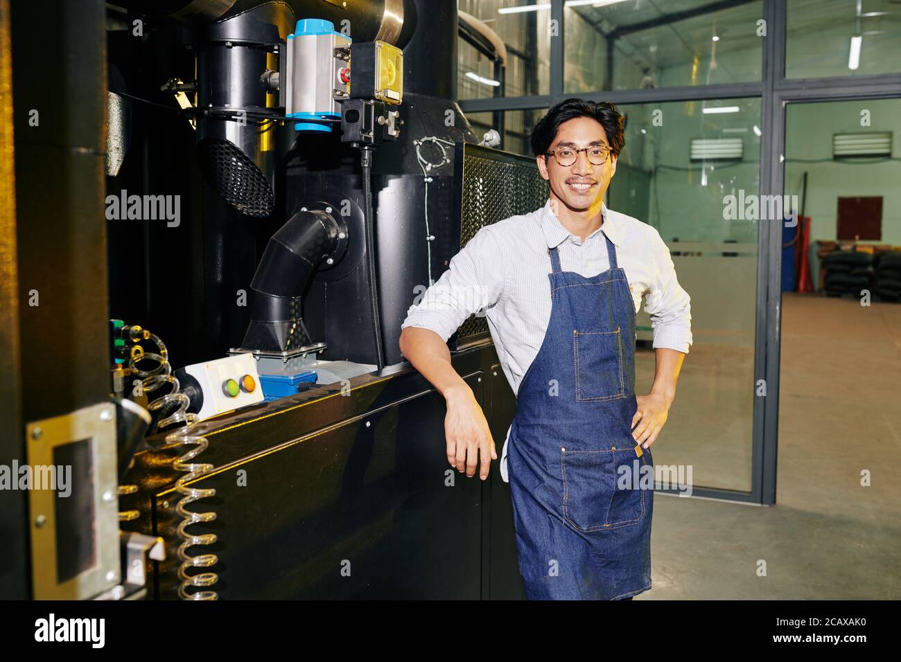 Portrait of positive barista in denim apron standing at big modern ...