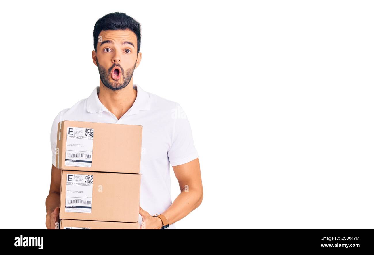 Young handsome hispanic man holding delivery package scared and amazed ...