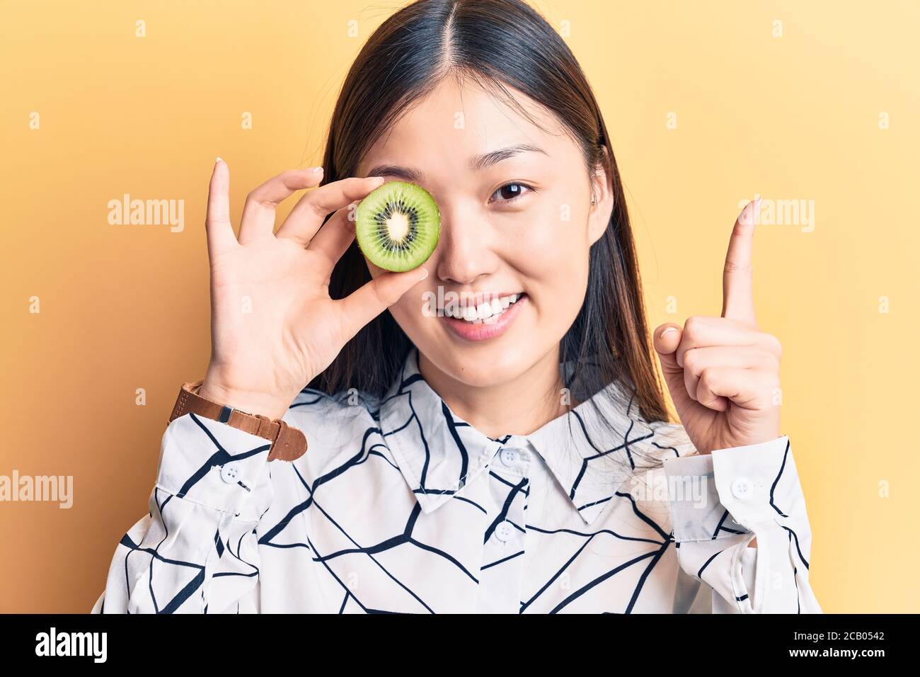 Young beautiful chinese woman holding kiwi over eye smiling with an ...