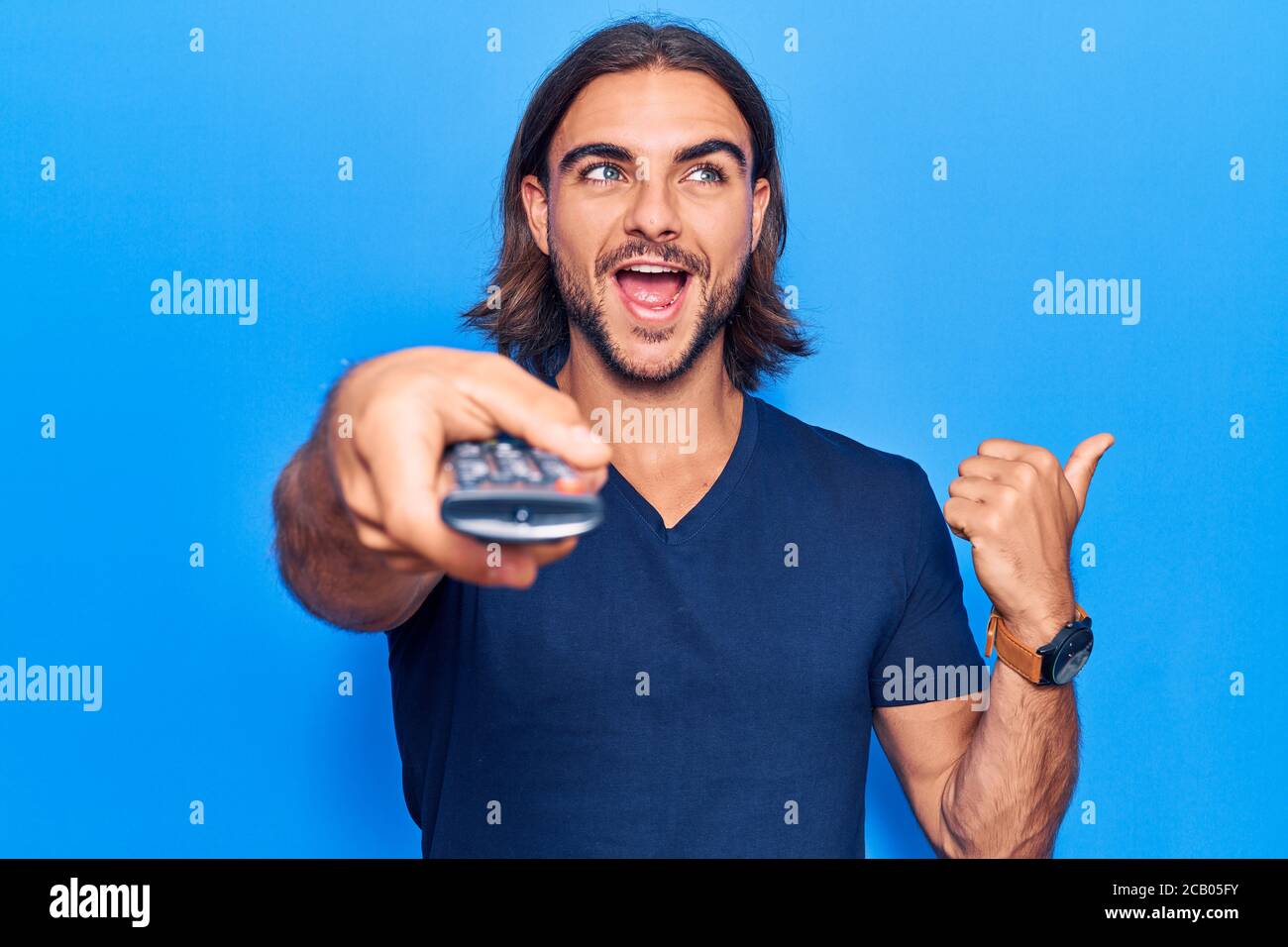 Young handsome man holding television remote control pointing thumb up ...