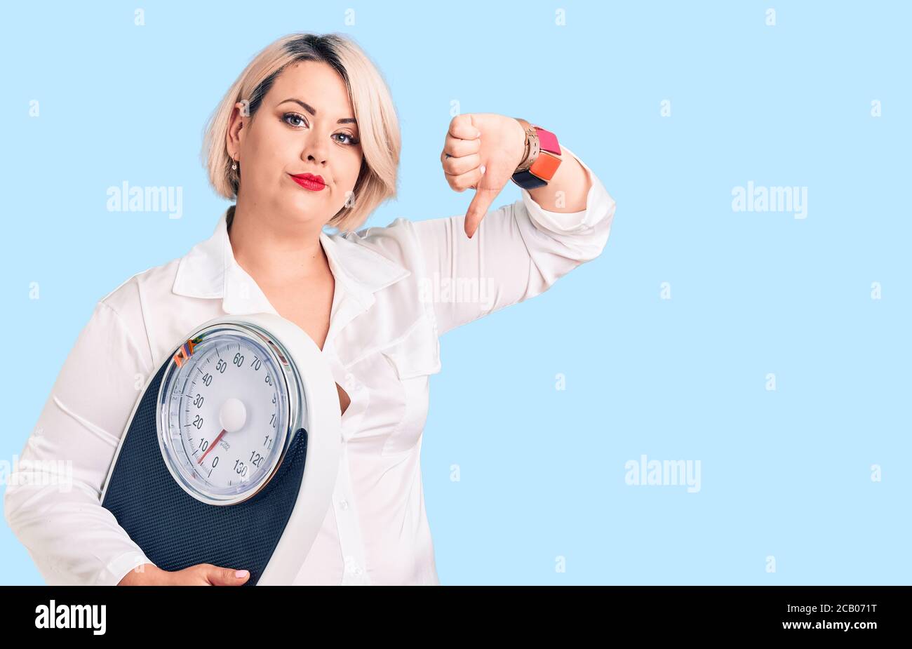 Young blonde plus size woman holding weighing machine with angry face ...