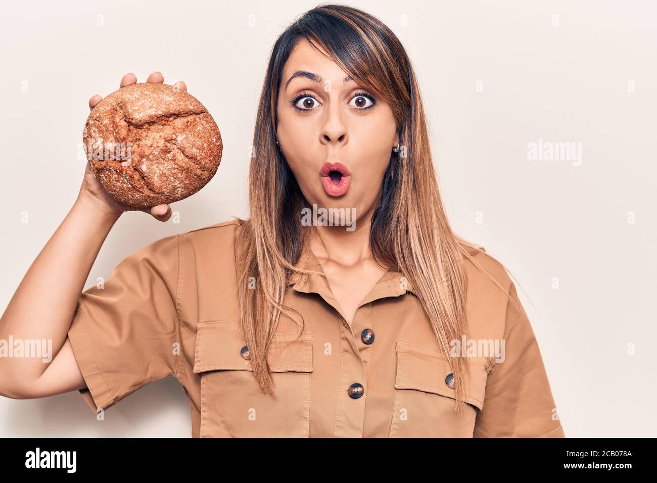 Young beautiful woman holding bread scared and amazed with open mouth ...
