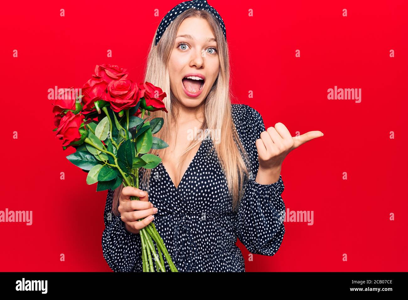 Young blonde woman holding flowers pointing thumb up to the side ...