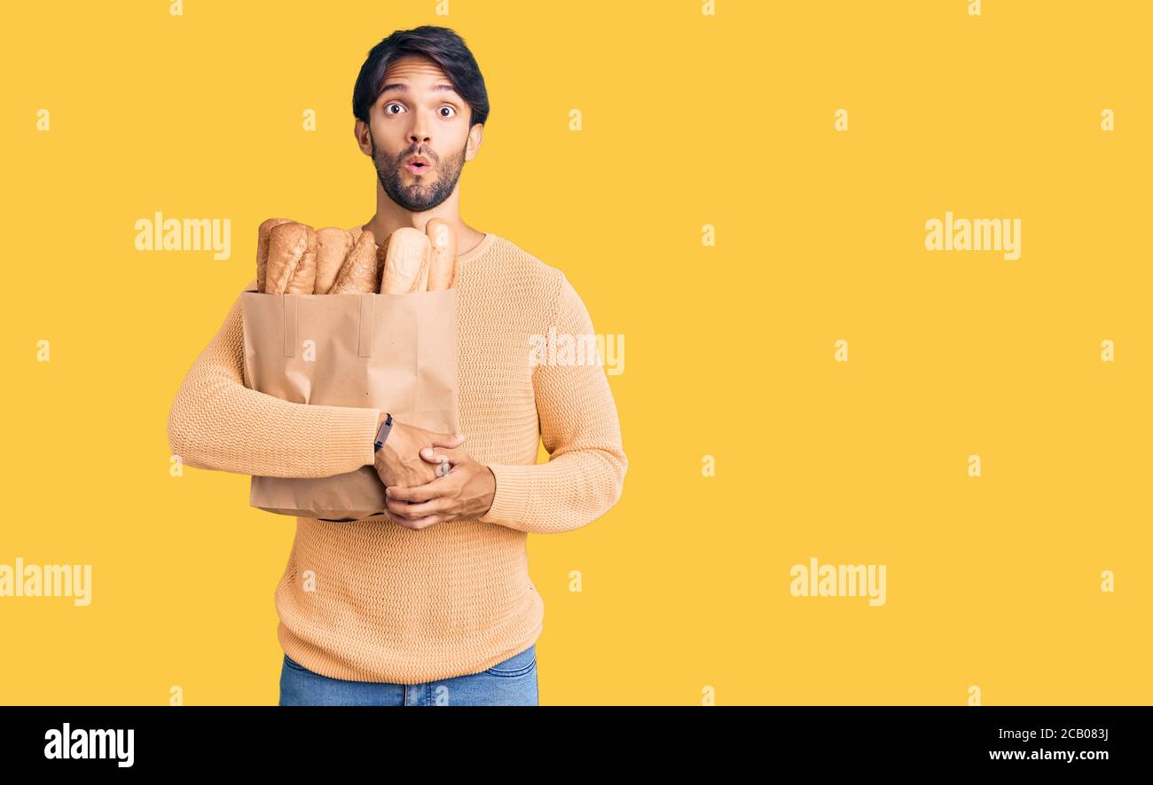 Handsome hispanic man holding paper bag with bread scared and amazed ...