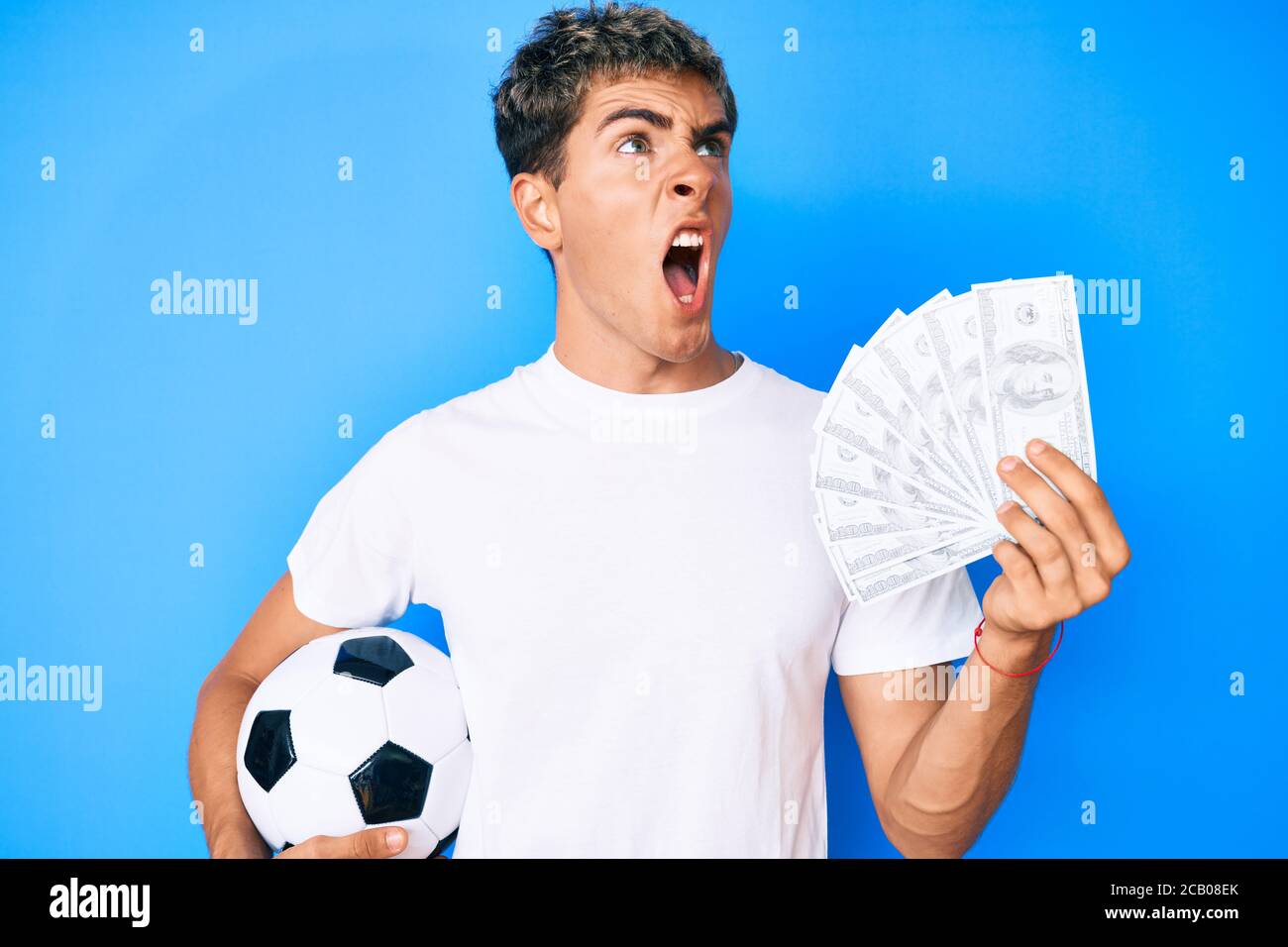 Young handsome man holding soccer ball and dollars banknotes angry and ...