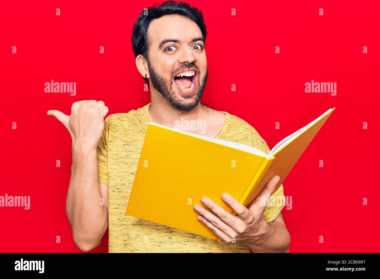 Young hispanic man holding book pointing thumb up to the side smiling ...