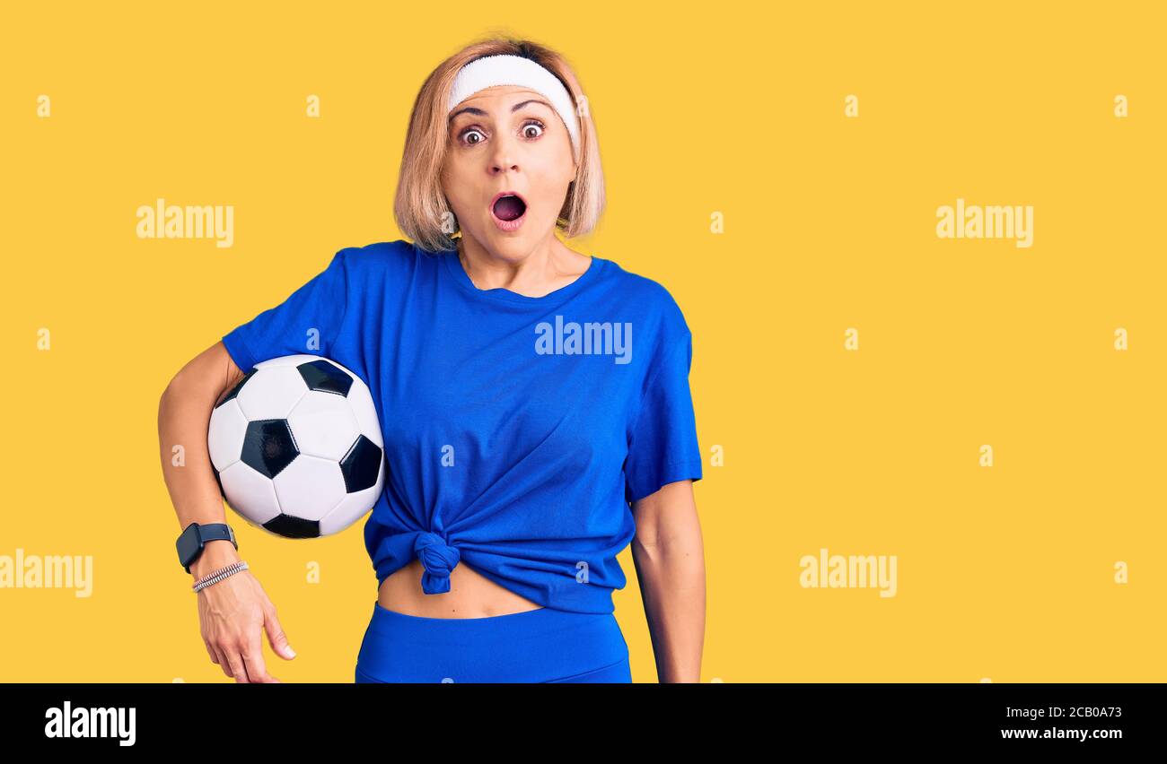 Young blonde woman holding soccer ball scared and amazed with open ...