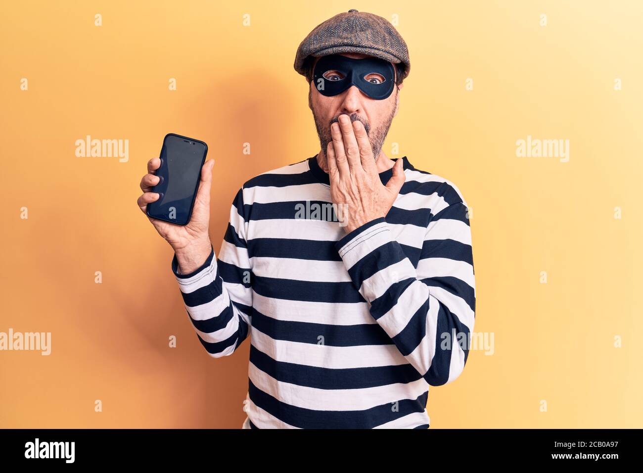 Middle age handsome burglar man wearing cap and mask holding smartphone ...