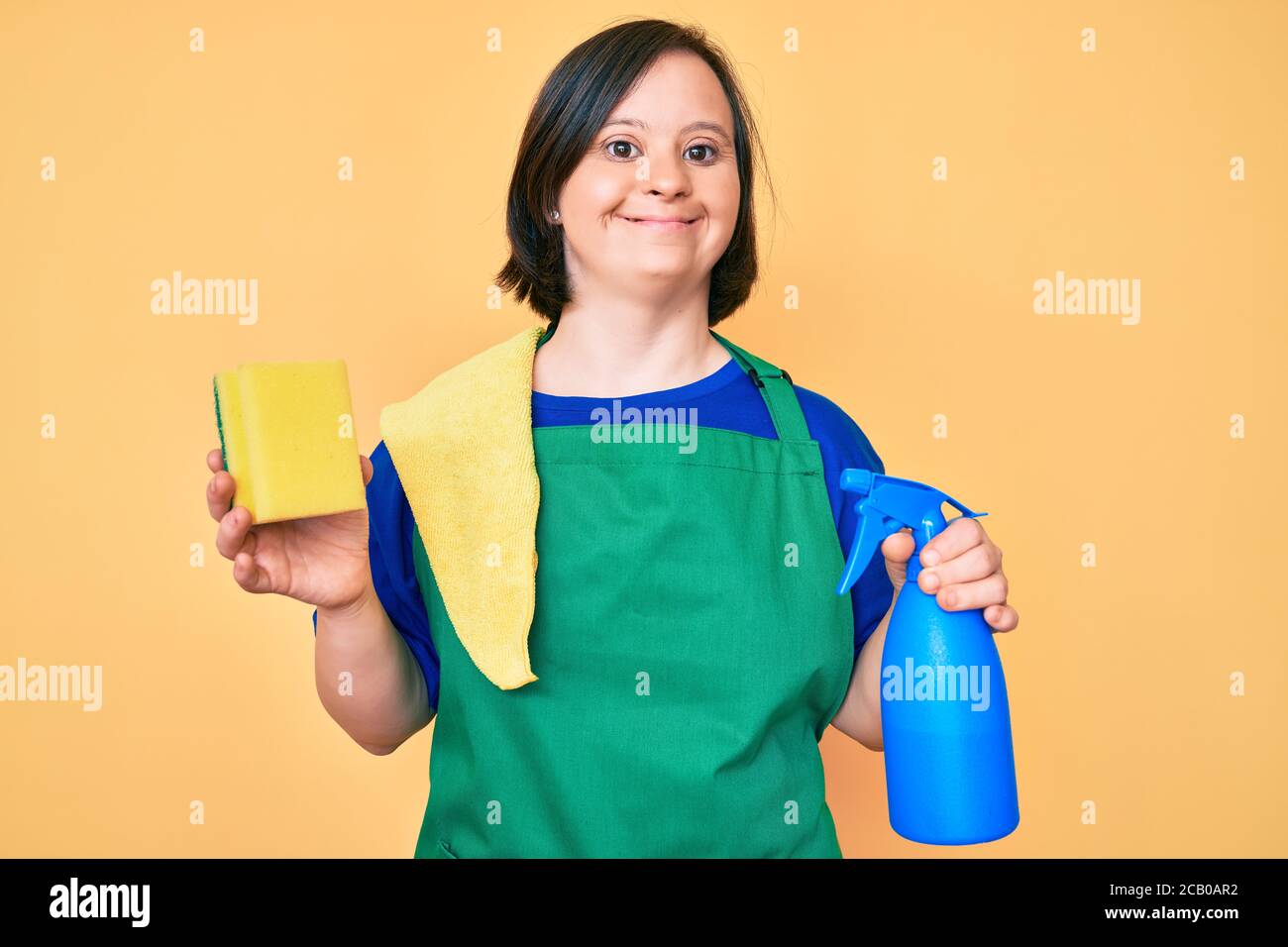 Brunette woman with down syndrome wearing apron holding scourer smiling ...