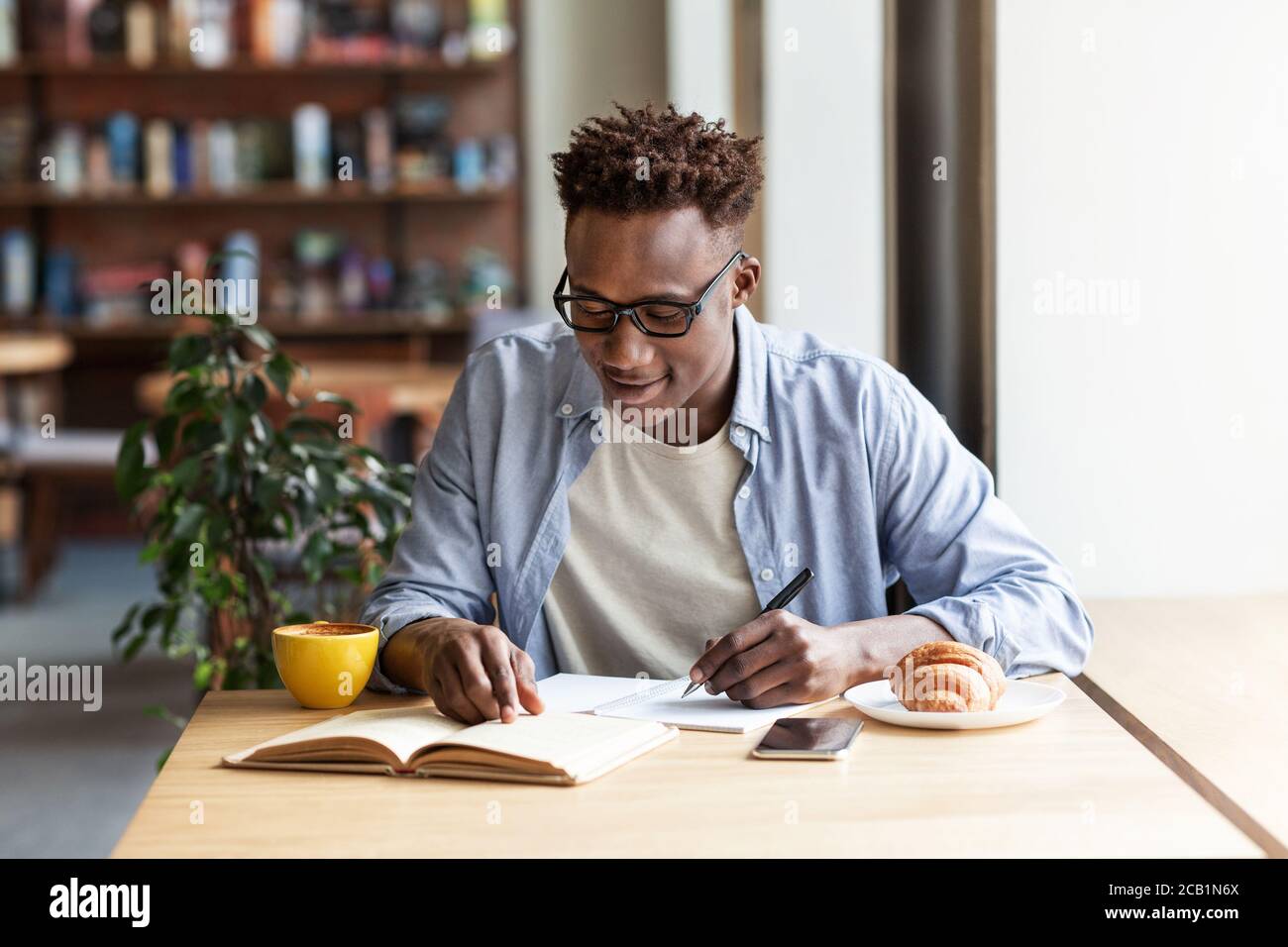 Focused male student taking notes from textbook at table in coffee shop ...