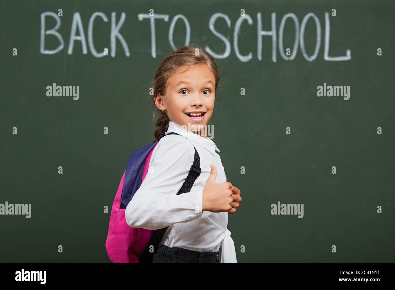 excited schoolgirl showing thumb up and looking at camera near ...