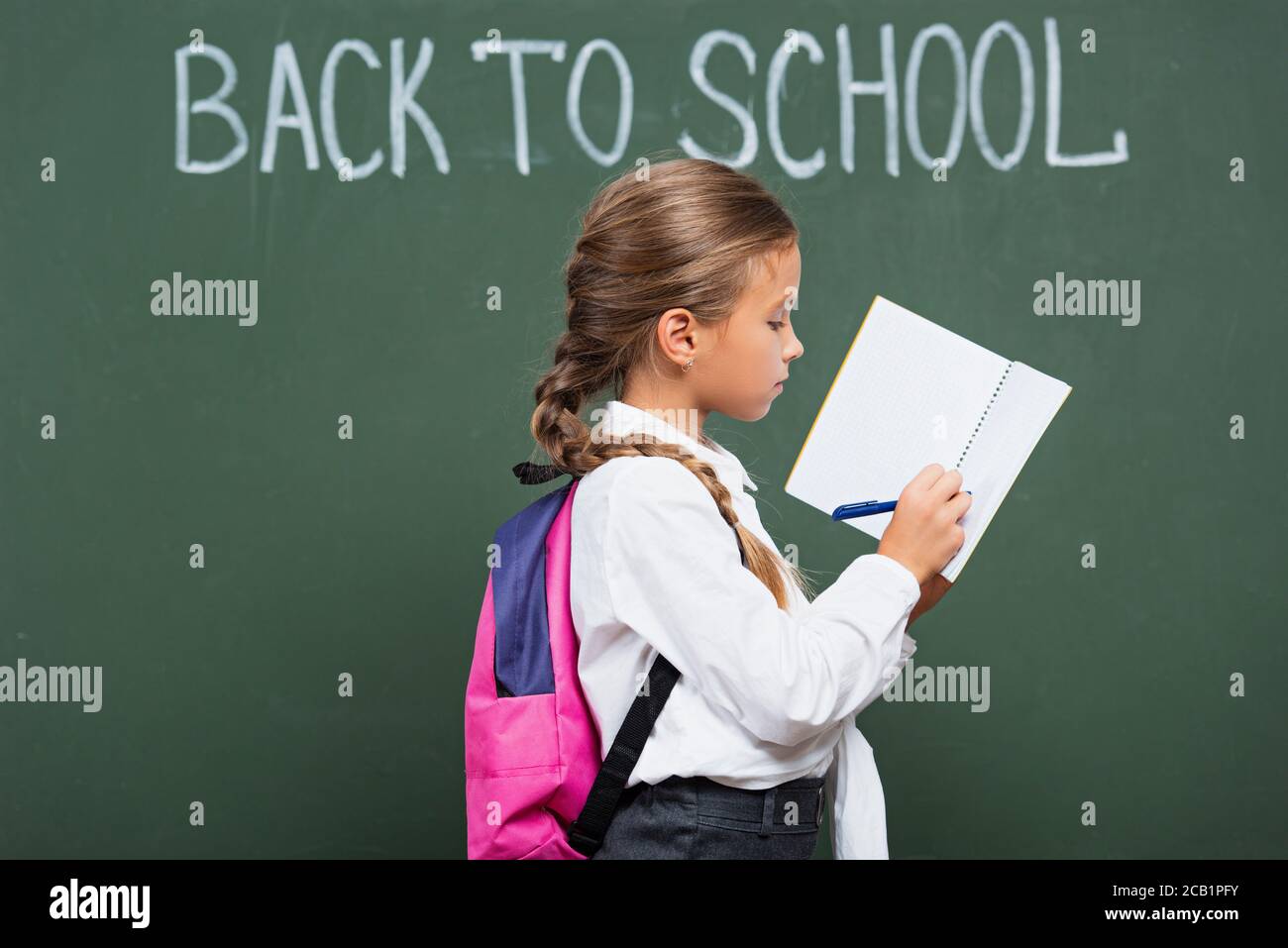schoolgirl with backpack writing in copy book near chalkboard with back ...
