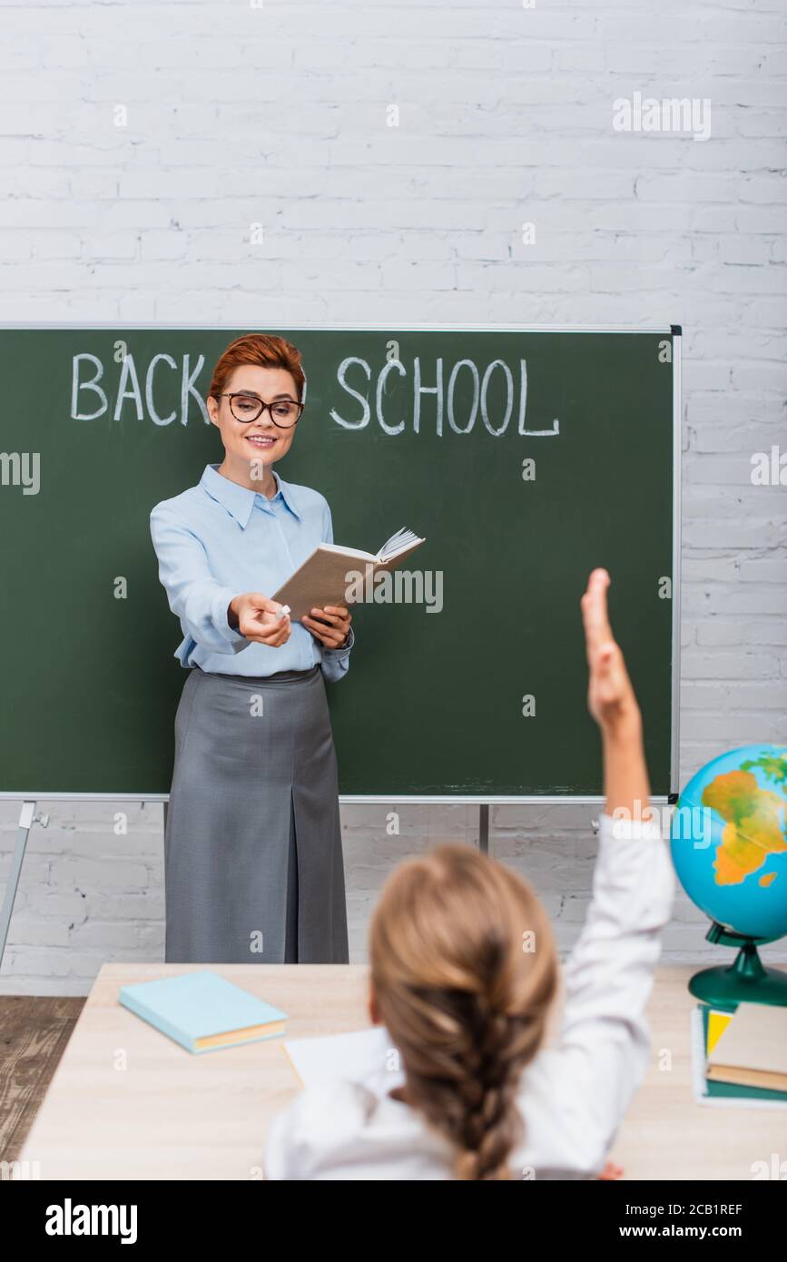 back view of schoolgirl with raised hand and smiling teacher near ...