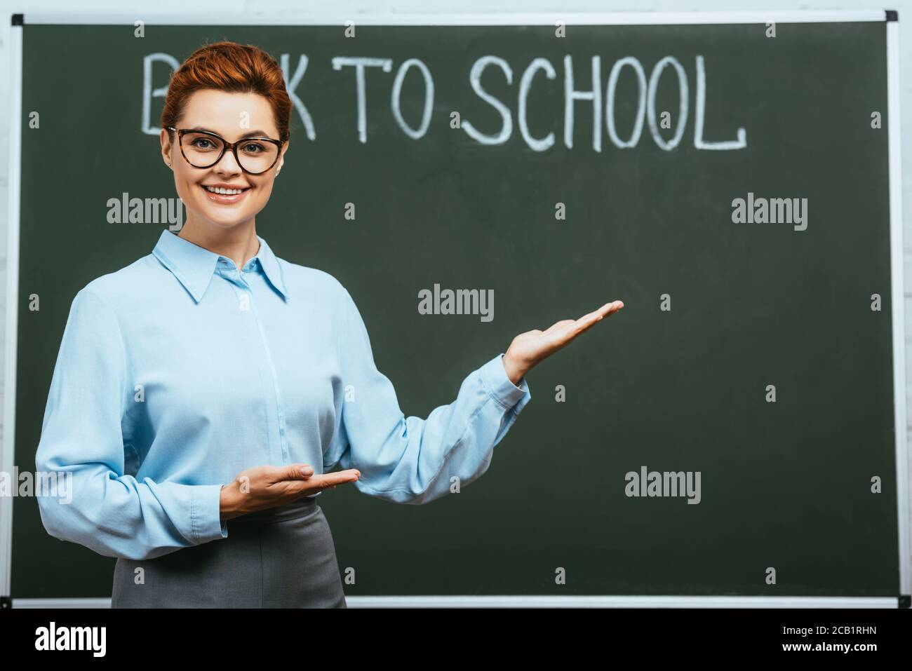 smiling teacher pointing with hands near chalkboard with back to school ...