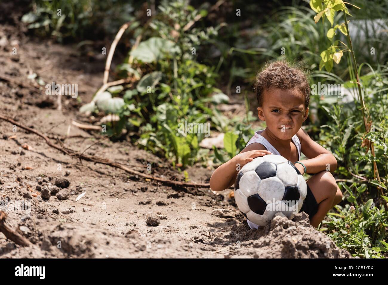 Poor african american boy holding football while sitting near plants on ...