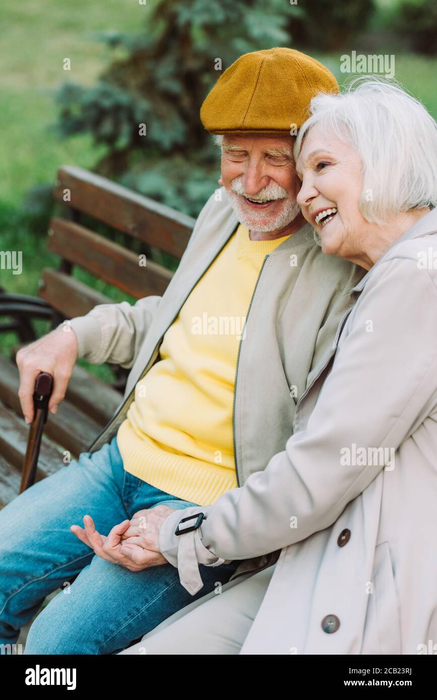 Smiling Elderly Couple Holding Hands While Sitting On Bench In Park