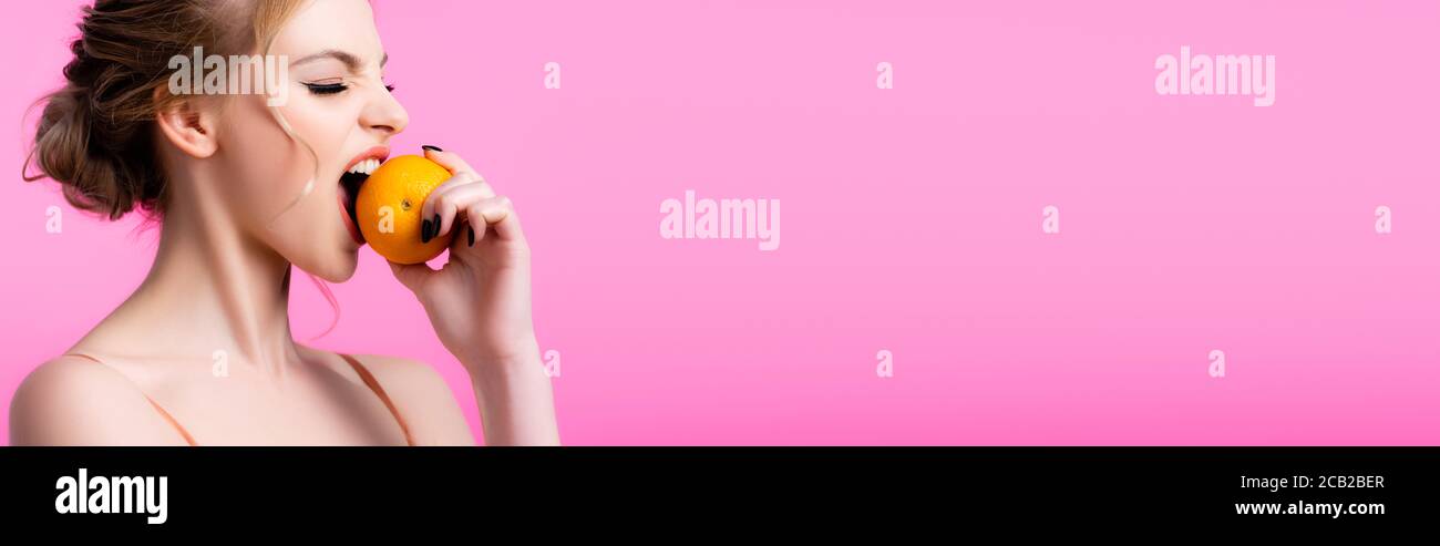 side view of beautiful blonde woman biting ripe orange isolated on pink ...