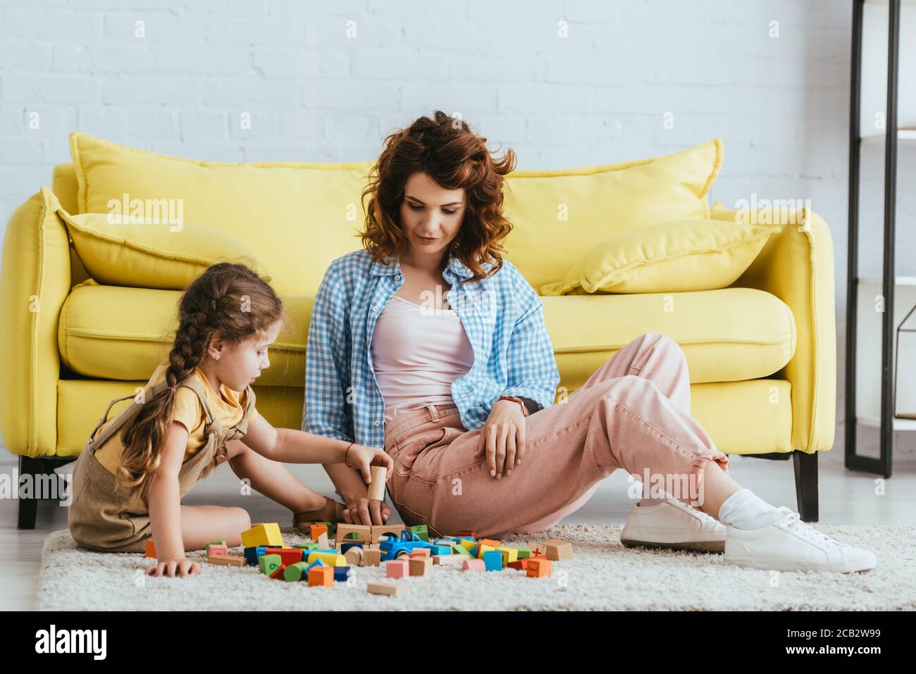 adorable kid holding block while playing on floor near young nanny ...