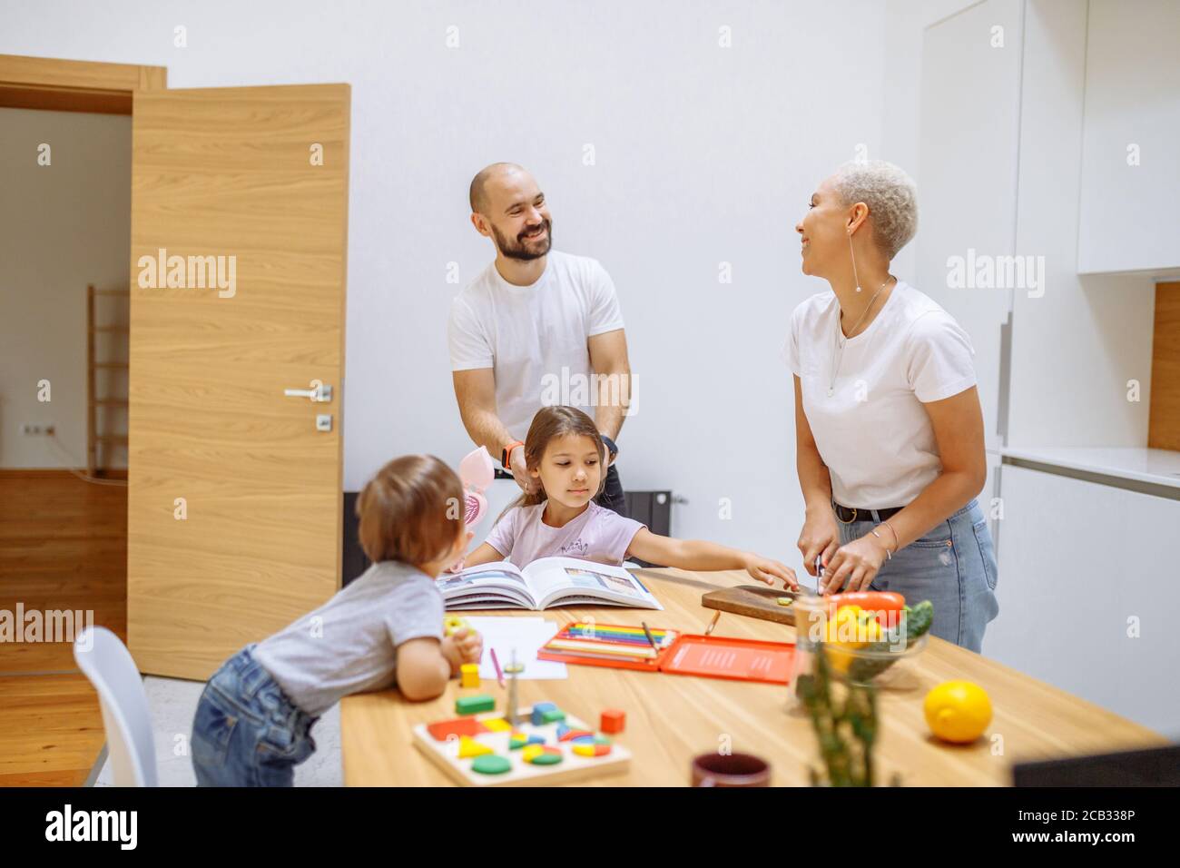 Parents teaching little girl homework, standing. Children sit at table ...