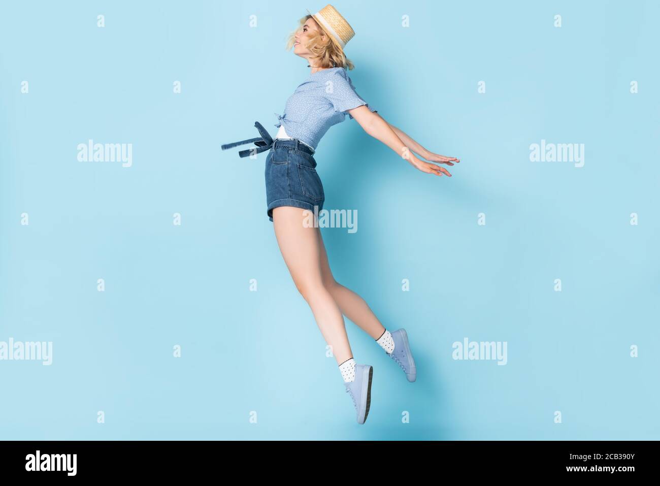 side view of young woman in shorts and straw hat jumping on blue Stock ...