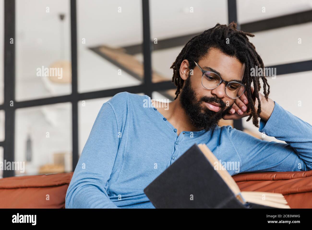 Handsome young african man sitting on a leather couch at home, reading ...