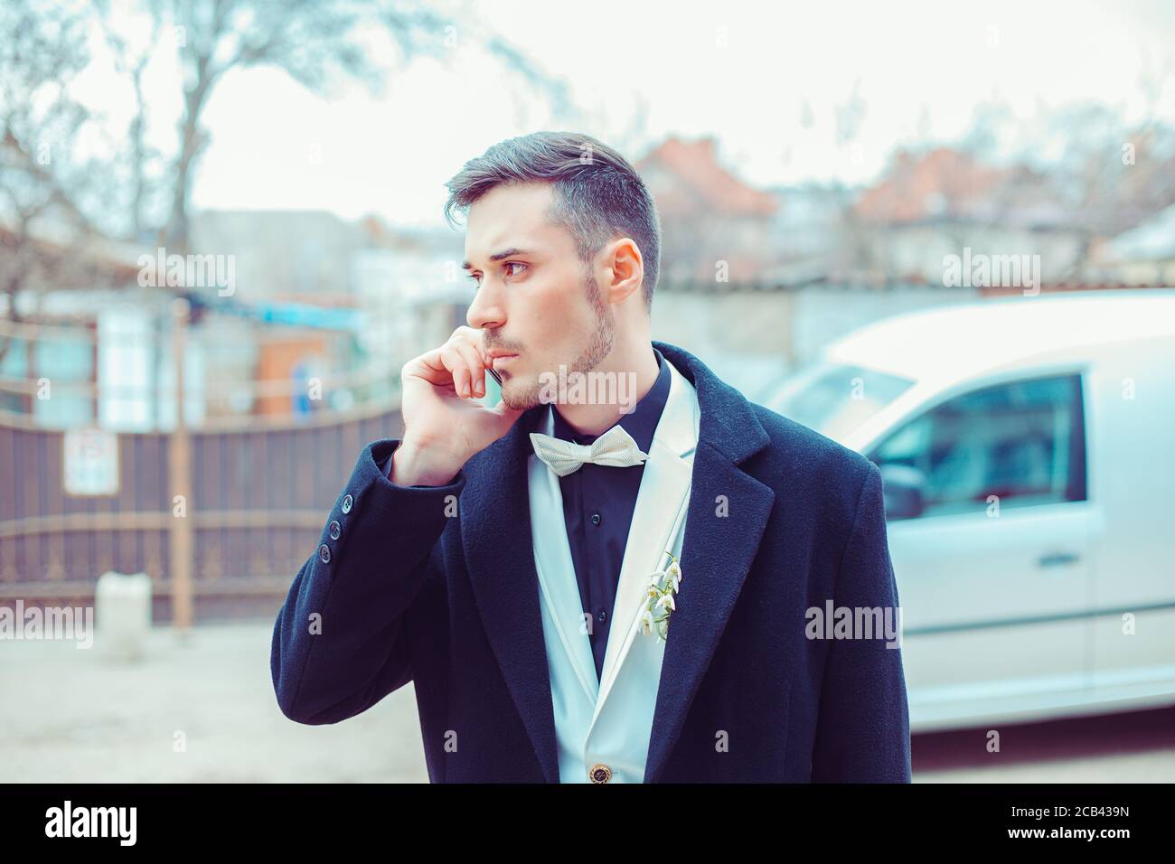 Trendy young man in wedding suit and coat having phone call standing on ...