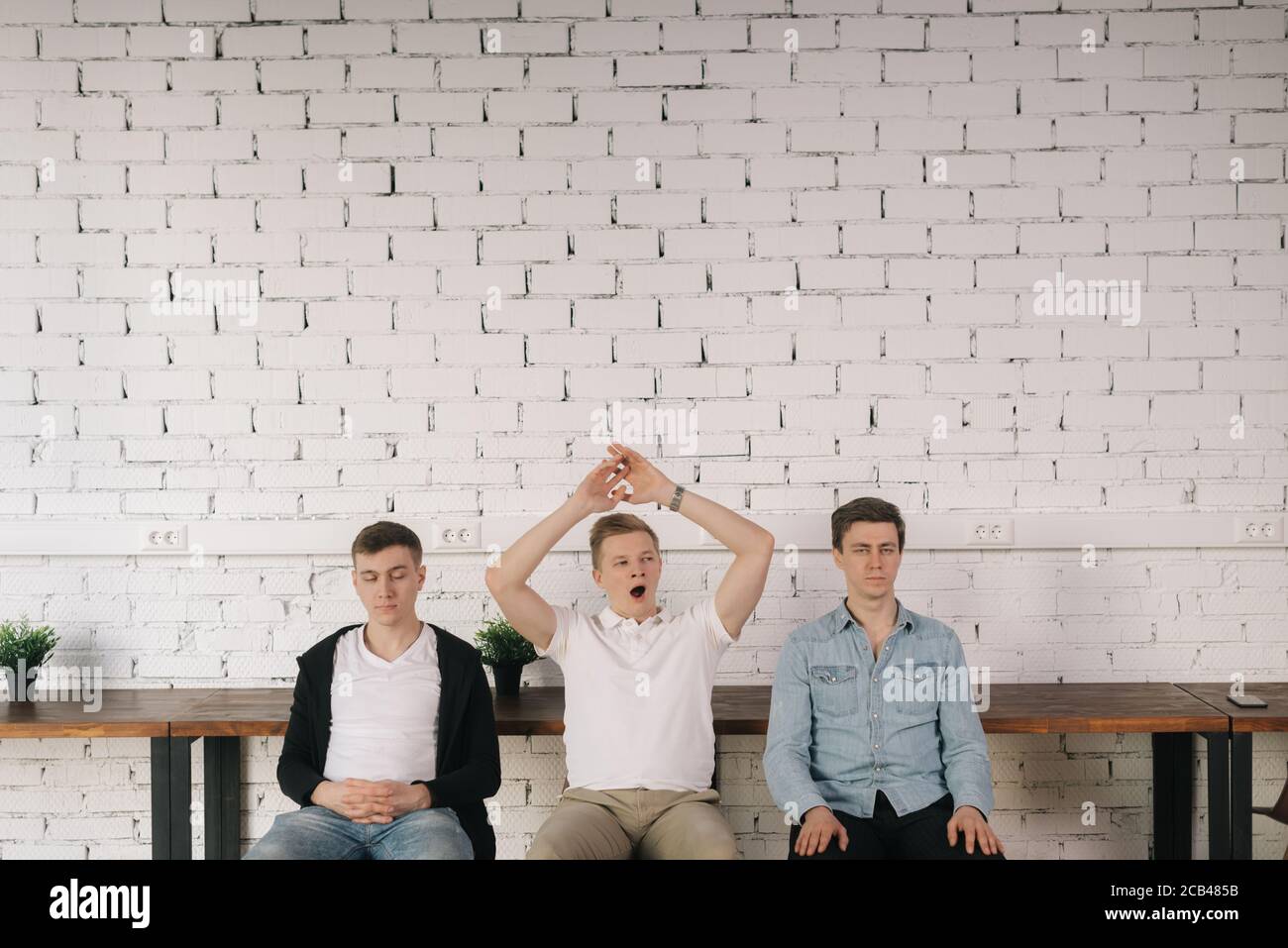 Three young tired guys sitting against a white brick wall waiting for ...